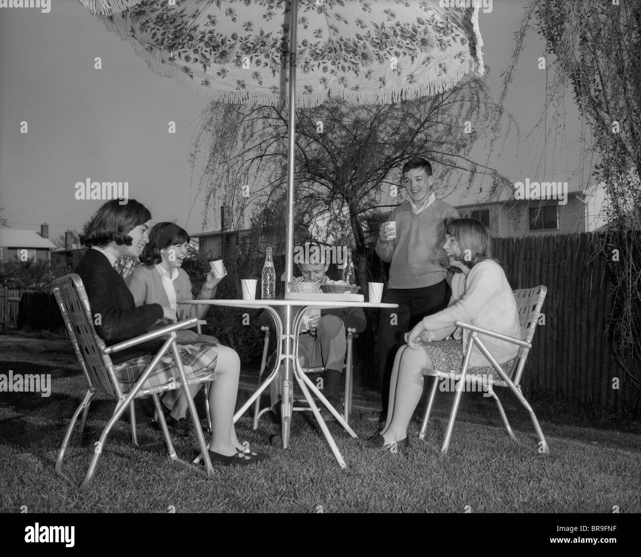 1960s GROUP TEENS SITTING IN BACKYARD UNDER LAWN UMBRELLA EATING SNACKS ...