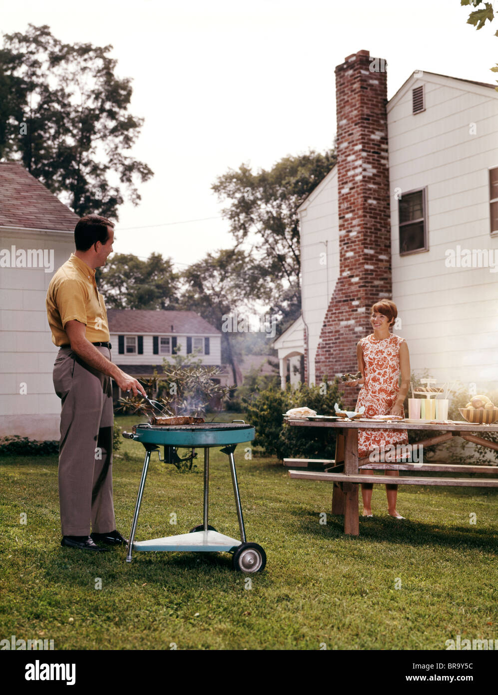 1960s COUPLE SUBURBAN BACKYARD MAN GRILLING WOMAN SETTING PICNIC TABLE ...