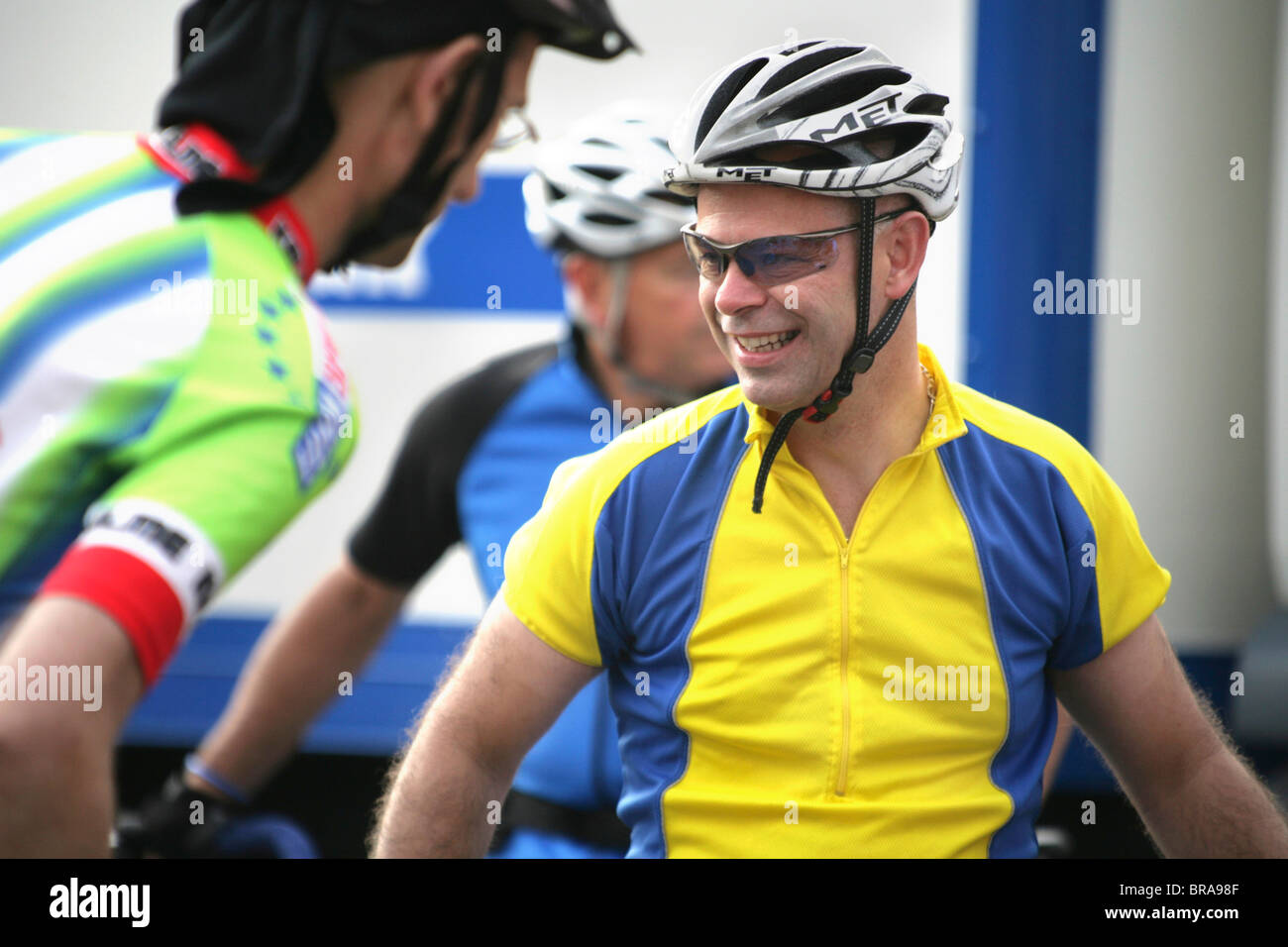Three men in cycling gear, wearing helmets. One man is smiling and ...