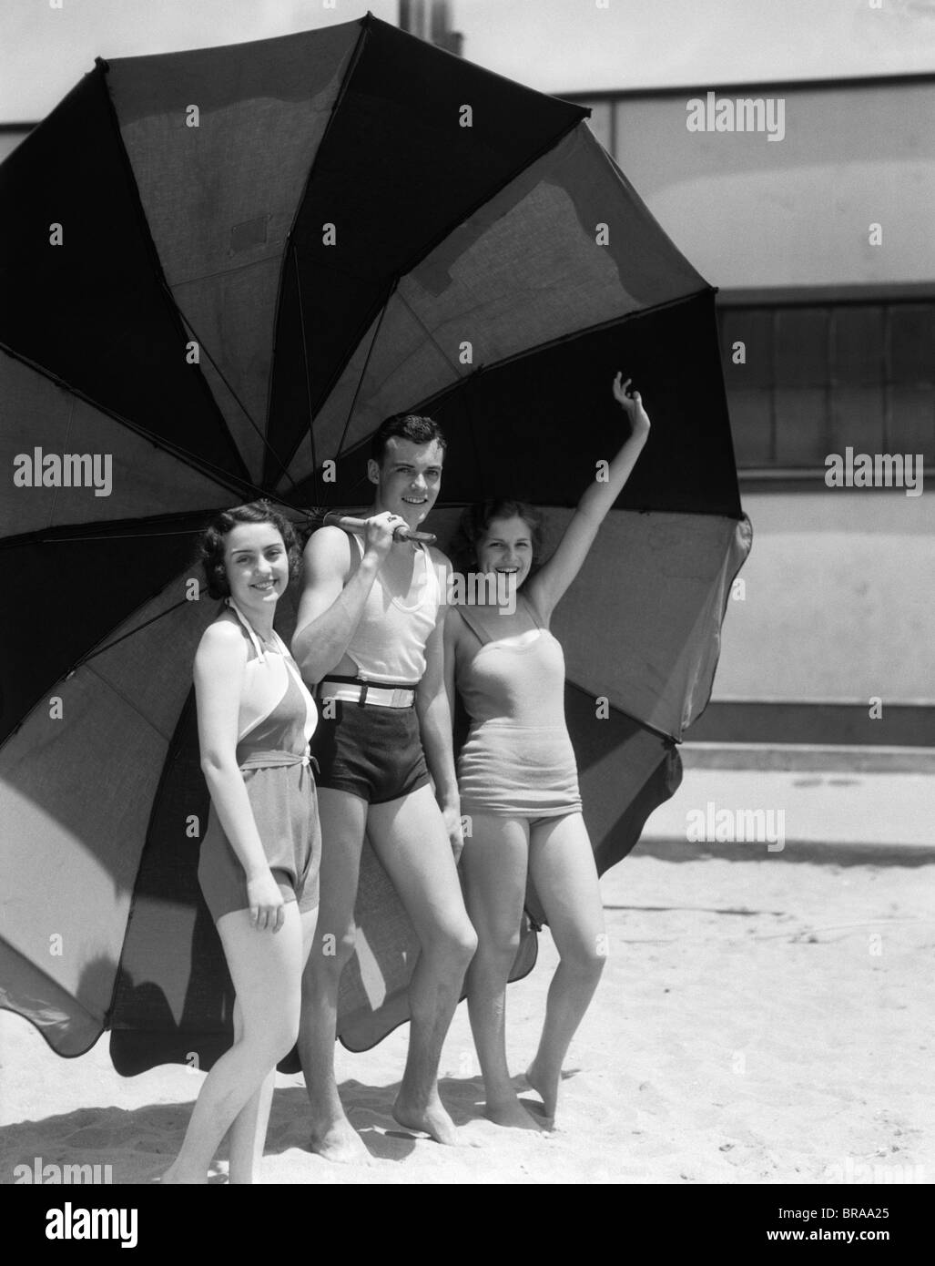 1930s TWO WOMEN ONE MAN SMILING WEARING BATHING SUITS STANDING UNDER EXTRA LARGE STRIPED BEACH ...