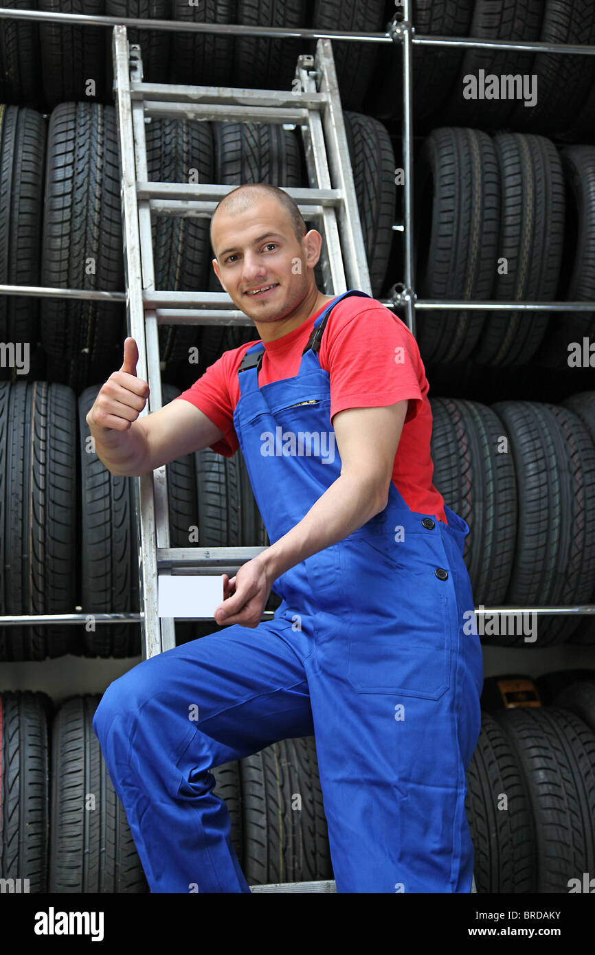 A smiling mechanic in a garage standing an a ladder next to a rack full of tires and making a ...