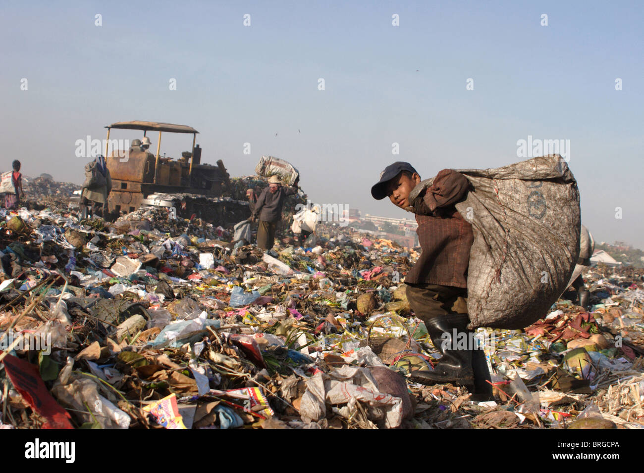 A young child laborer carries a big heavy bag of garbage while working ...