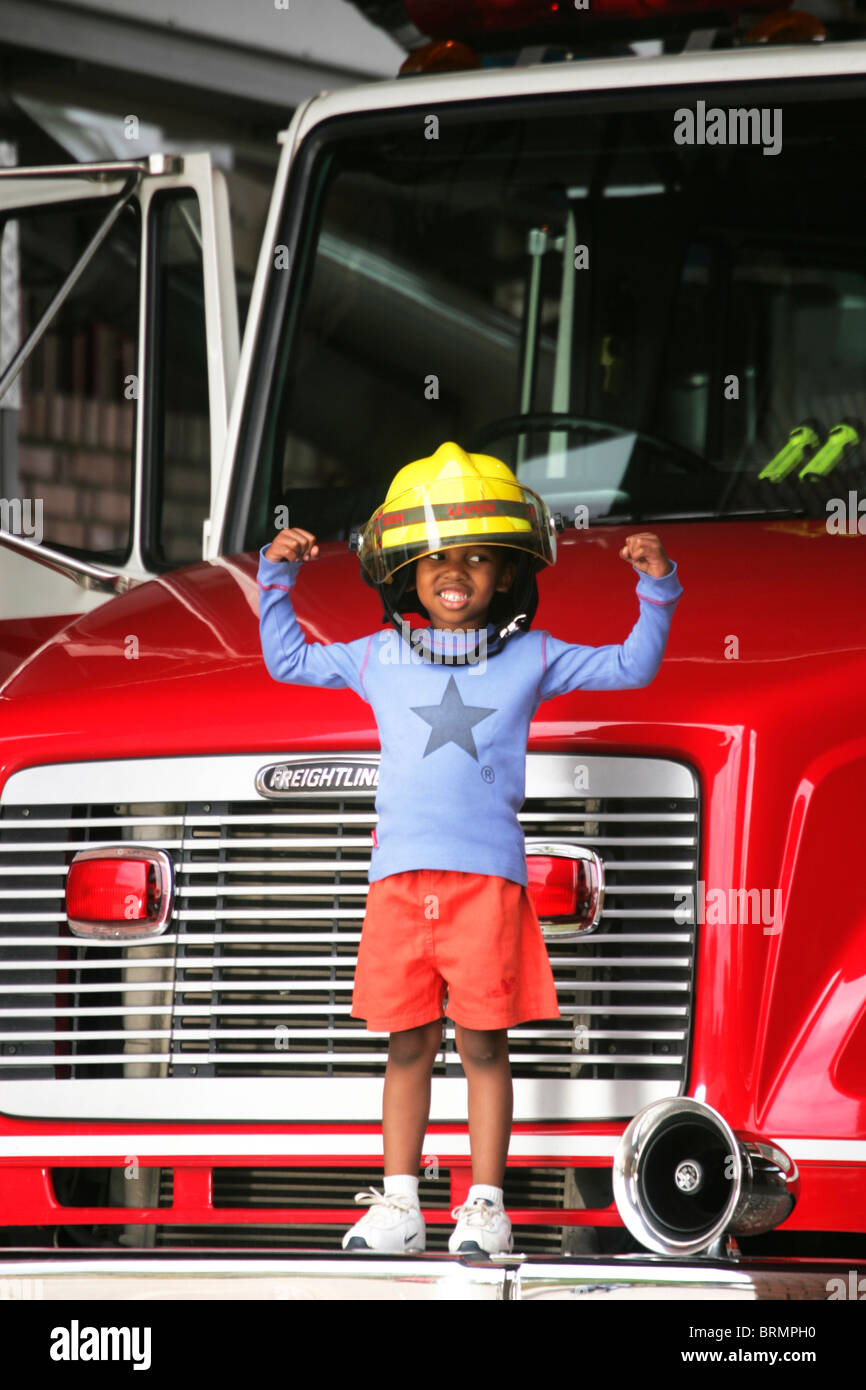 A young boy standing on the bumper of a fire engine wearing a fire ...
