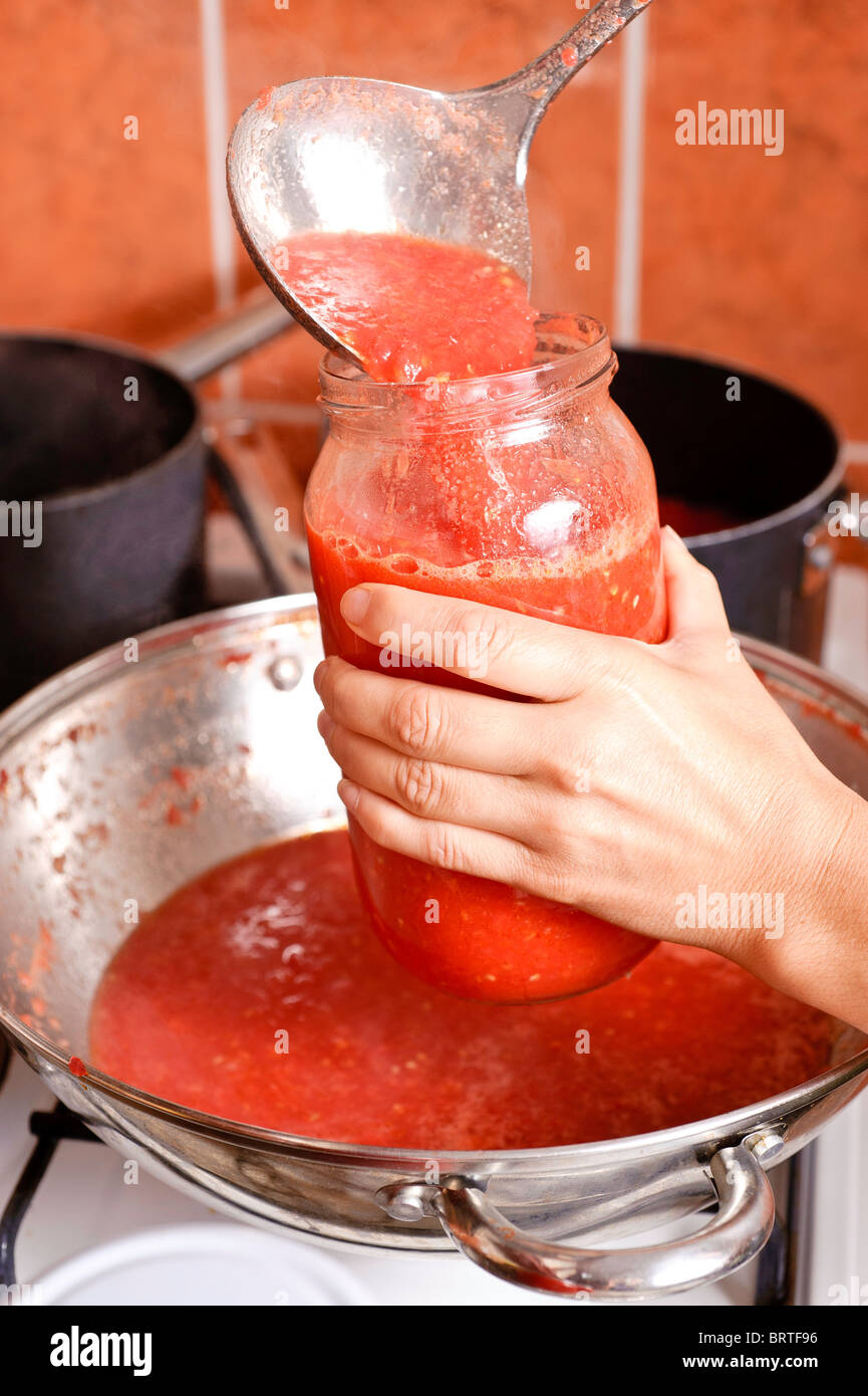 A woman's hand holds a half full glass jar whilst pouring heated tomato ...