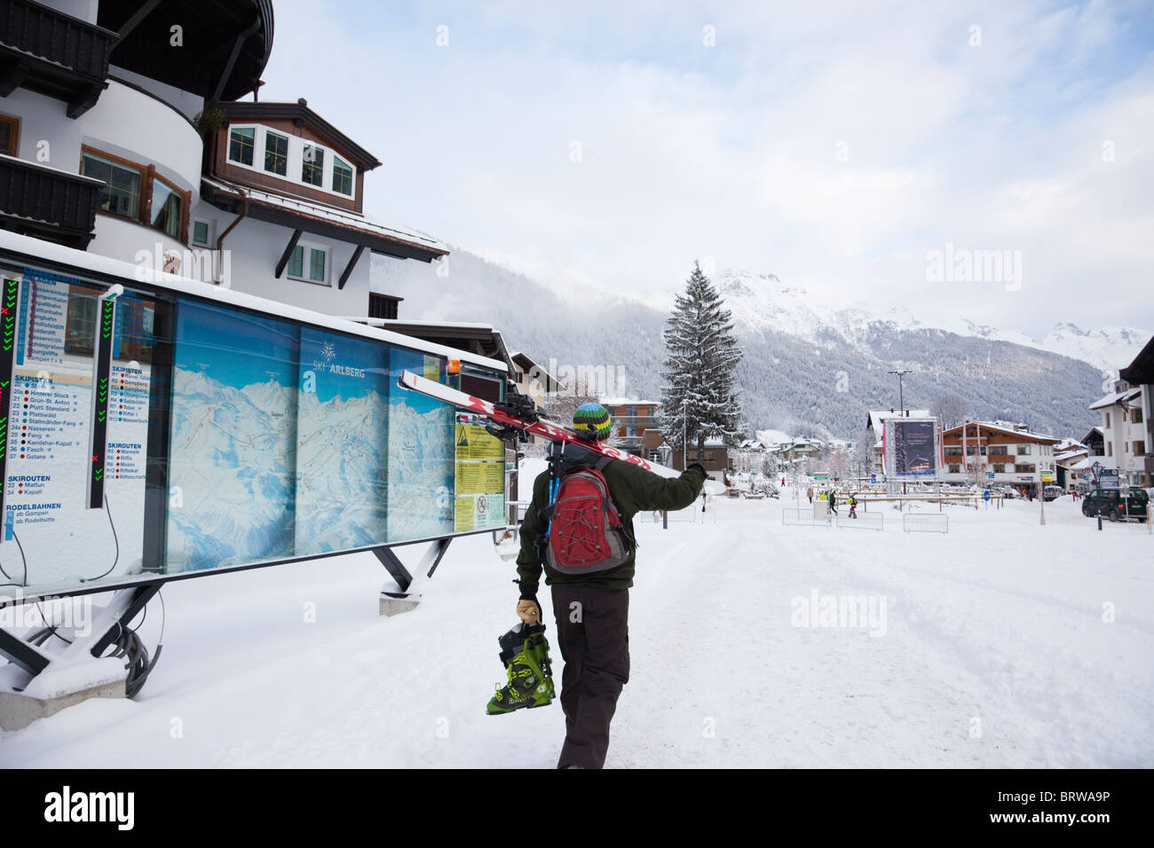 Skier carrying skis and ski boots passing piste information boards in ...