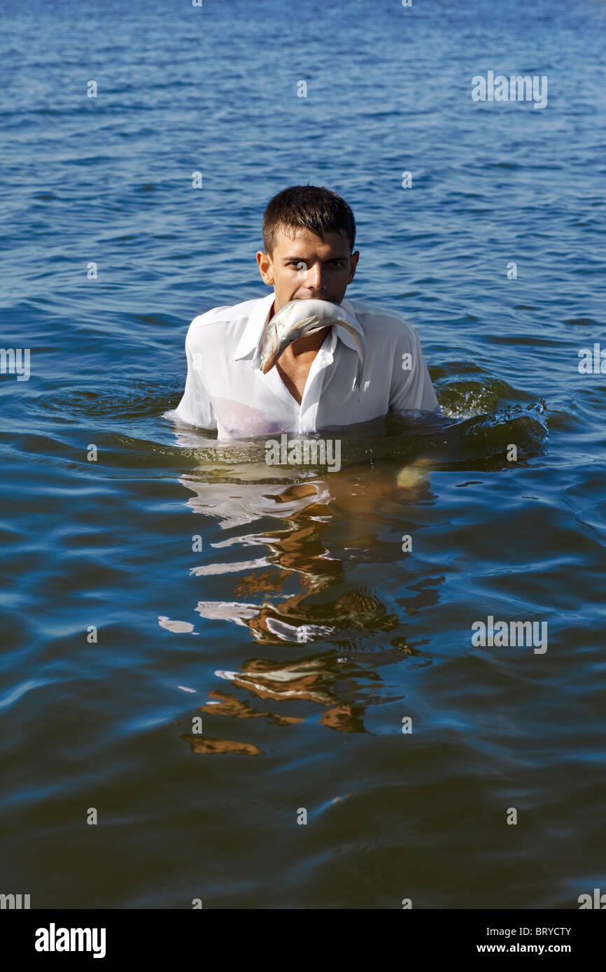 man in classic white shirt walking in water bearing fish in his mouth ...