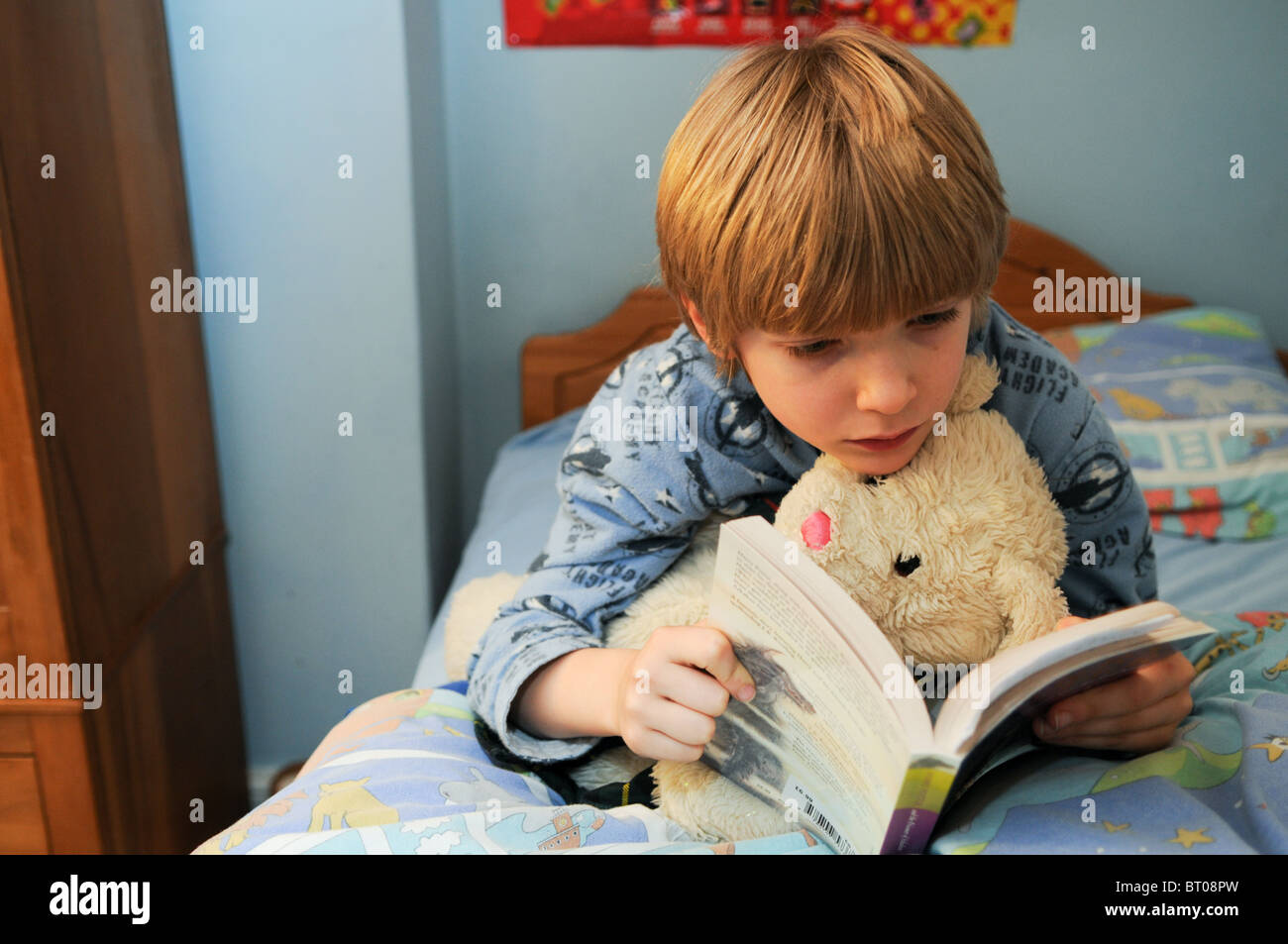 A young boy reading in bed wearing pajamas cuddling his teddy bear ...