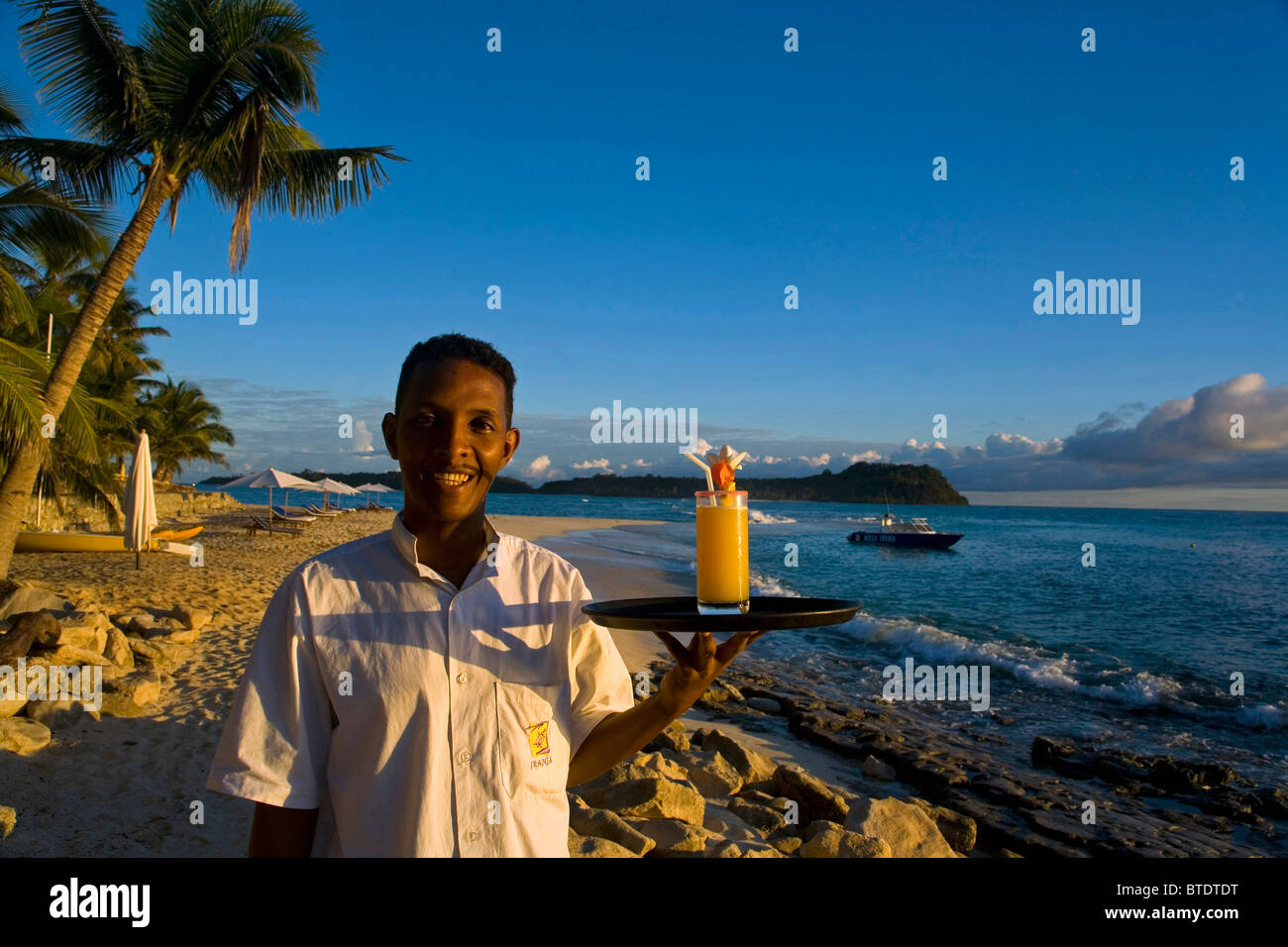 A smiling waiter standing on a beach balancing a tray with a glass of ...