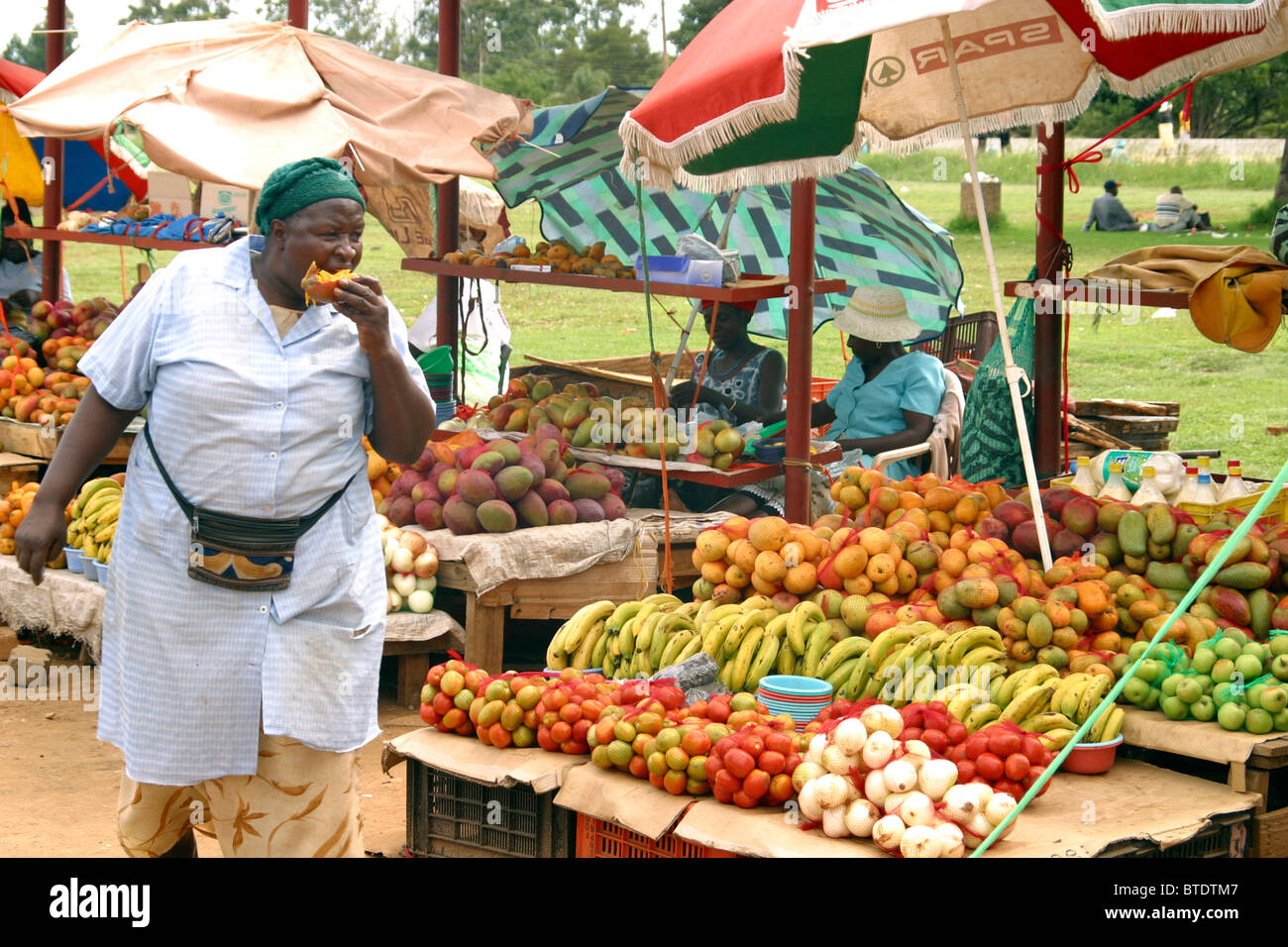 African woman vendor eating a mango at a roadside fruit and vegetable ...