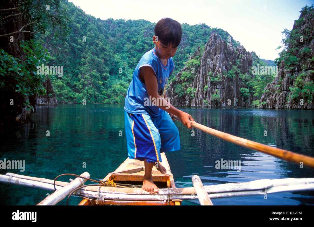 A boy navigating his small outrigger boat with a bamboo pole in shallow ...