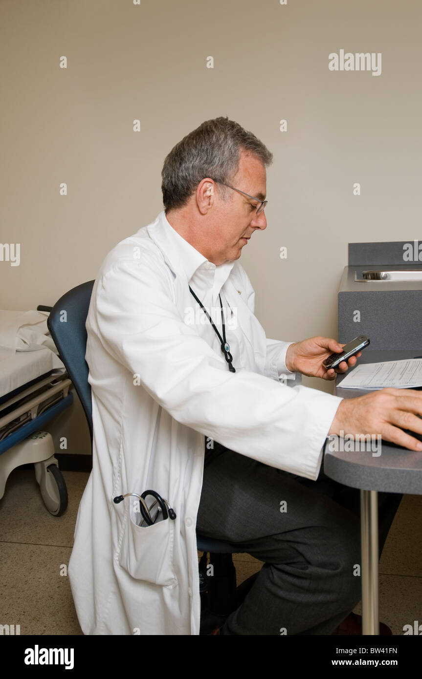 Doctor sitting at desk using mobile device in hospital examination room ...