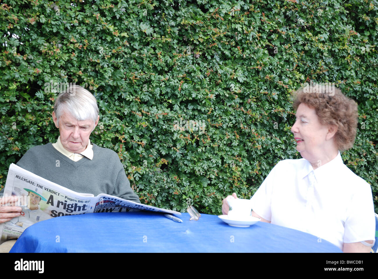 OAP couple sitting outdoors in their garden drinking tea and reading a ...