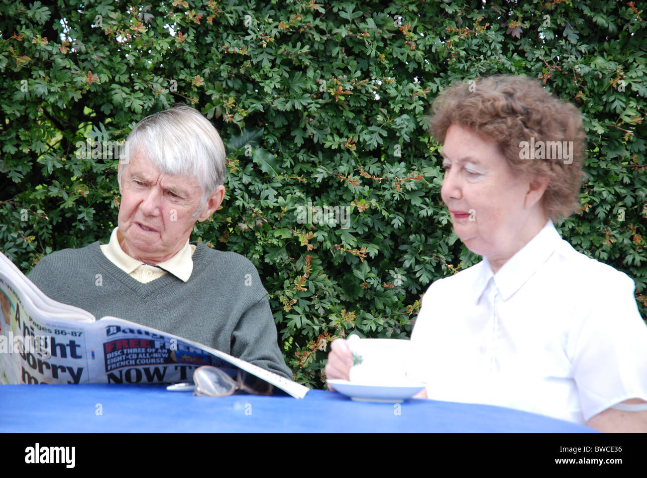 OAP couple sitting outdoors in their garden drinking tea and reading a ...