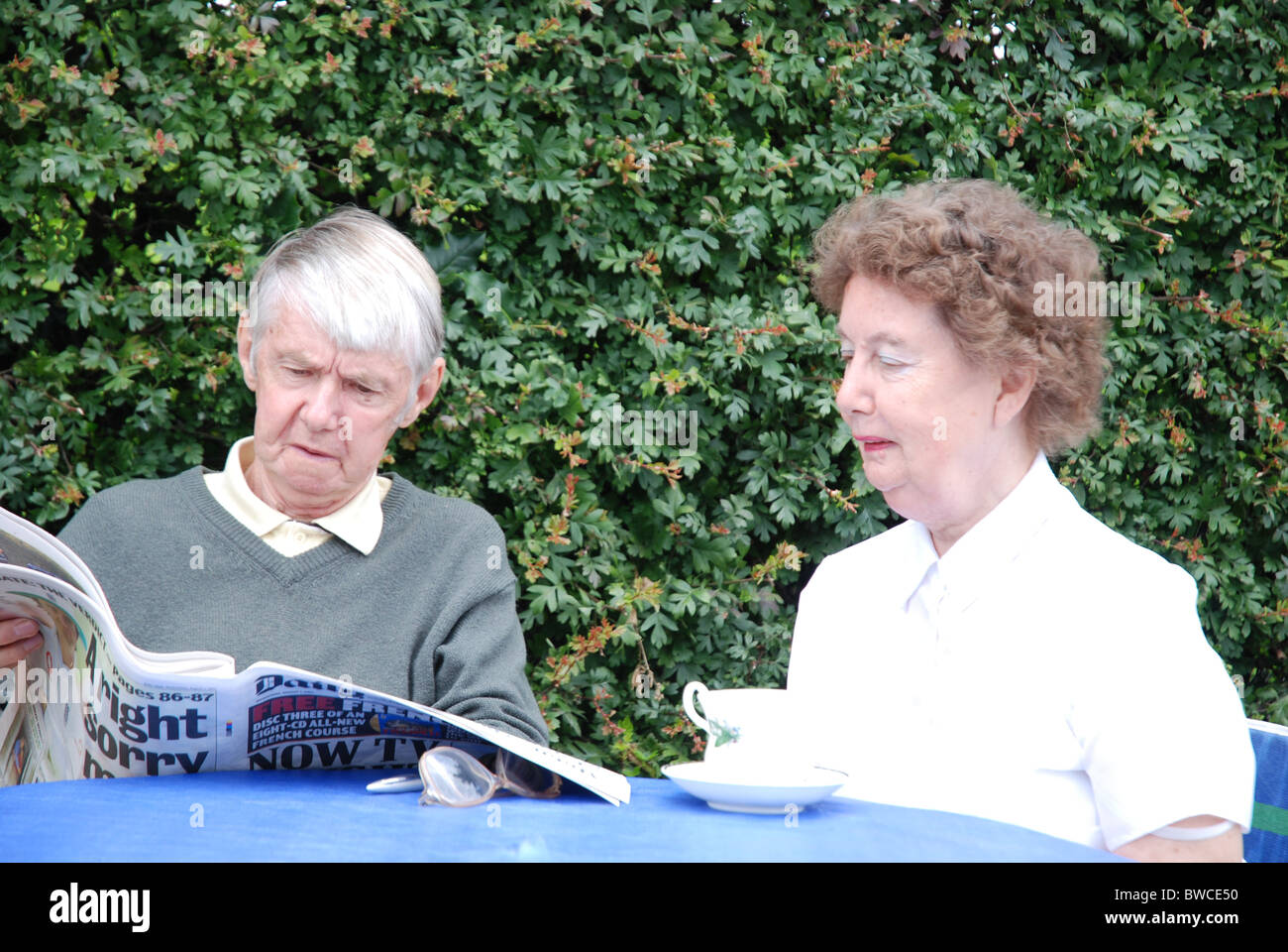 OAP couple sitting outdoors in their garden drinking tea and reading a ...