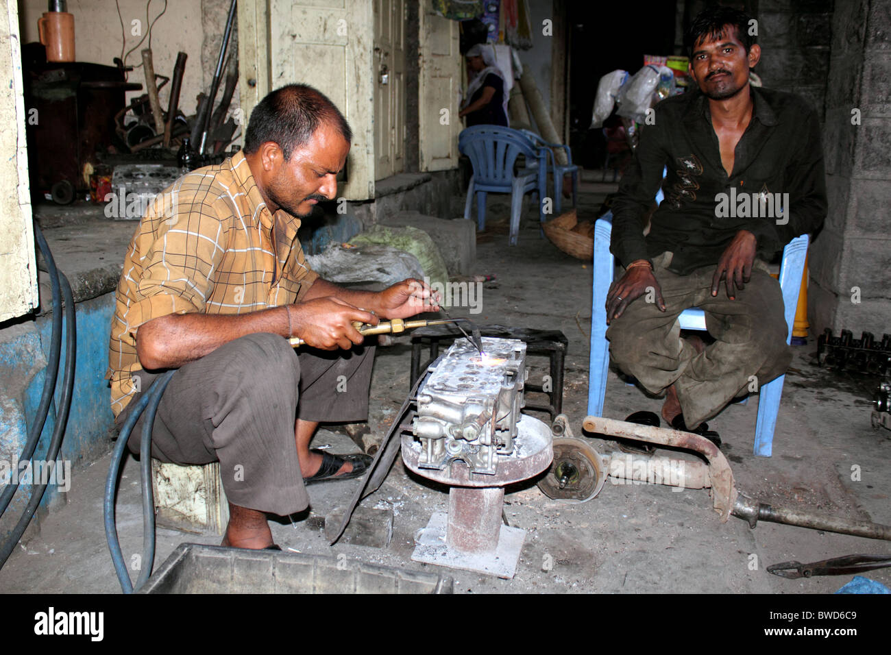 Indian man welds car engine block while another man sits and watches ...