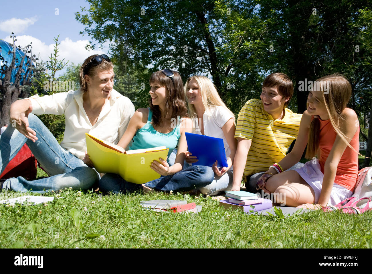 Portrait of cheerful friends talking and reading books in park together ...