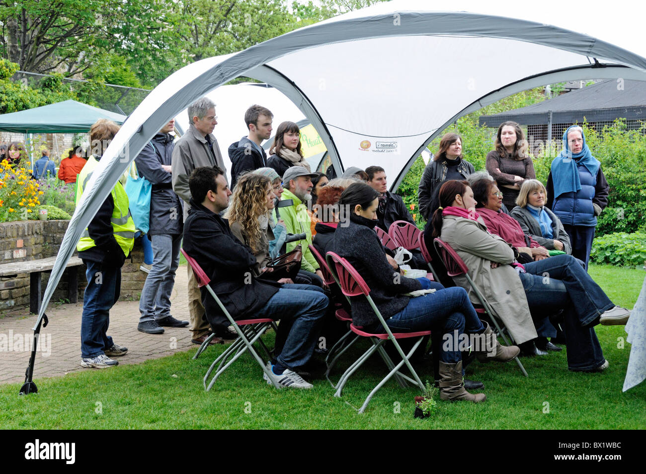 An audience of people sitting on chairs inside a small open marquee or ...