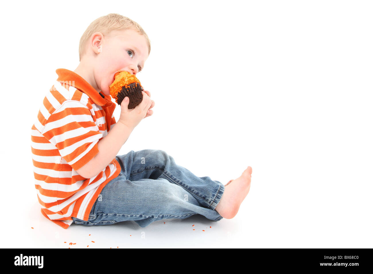 Adorable 2 year old boy eating orange icing cupcake over white ...