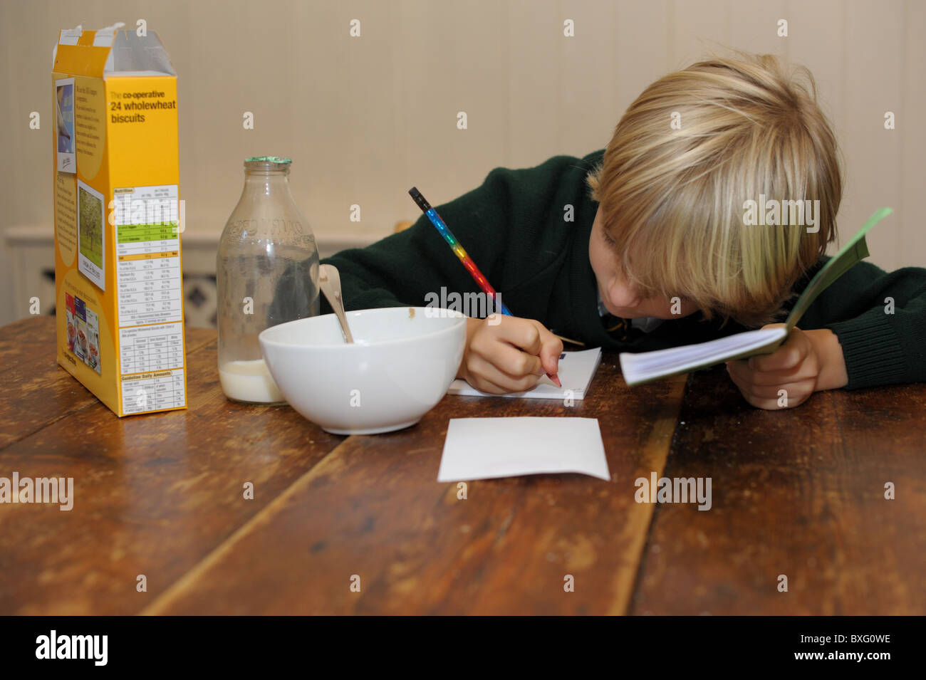 A young boy in his green school uniform sits at a wooden kitchen table ...