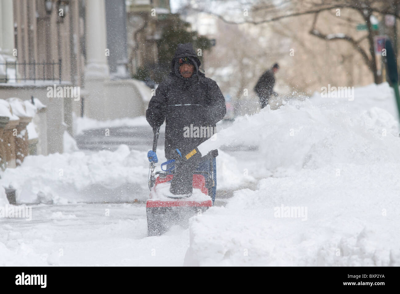 A man using a snow blower to clear the sidewalk in front of a building ...