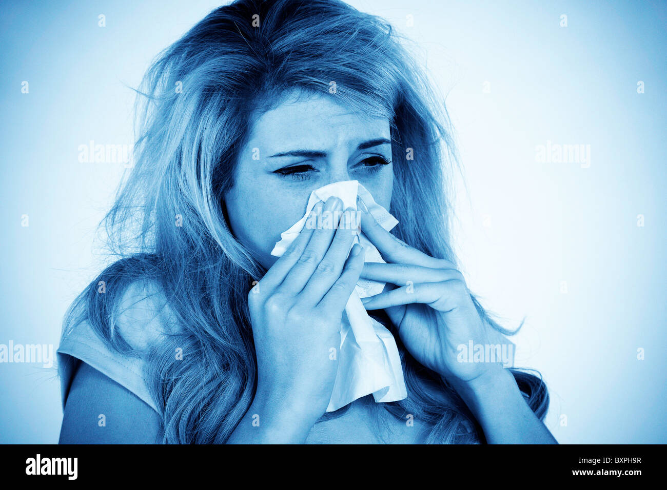 Young blond woman with a cold sneezing and blowing her nose on a tissue ...