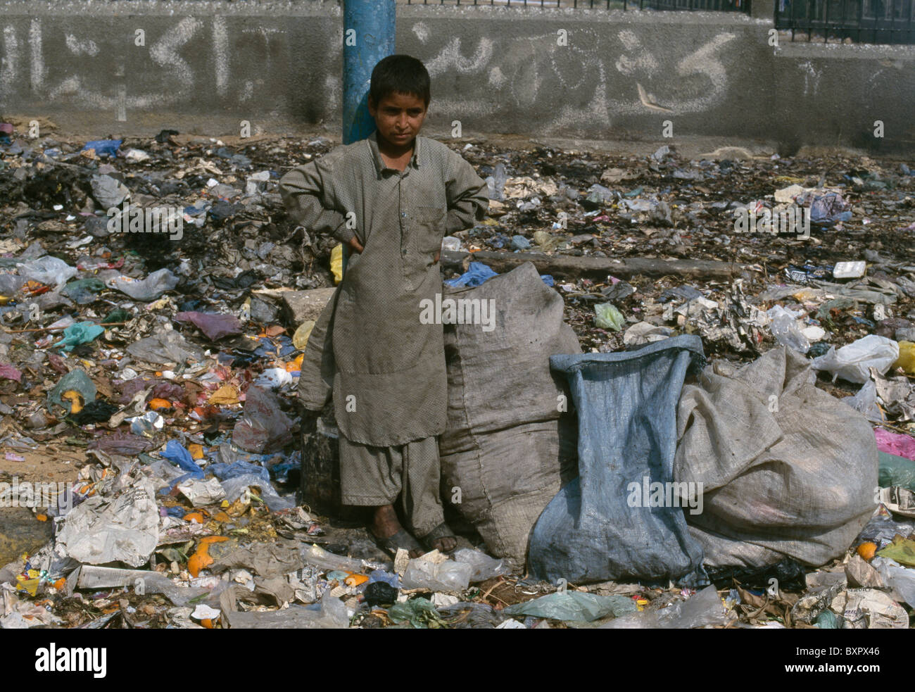 A young Pakistani boy stands in a garbage dump with his collection bags ...