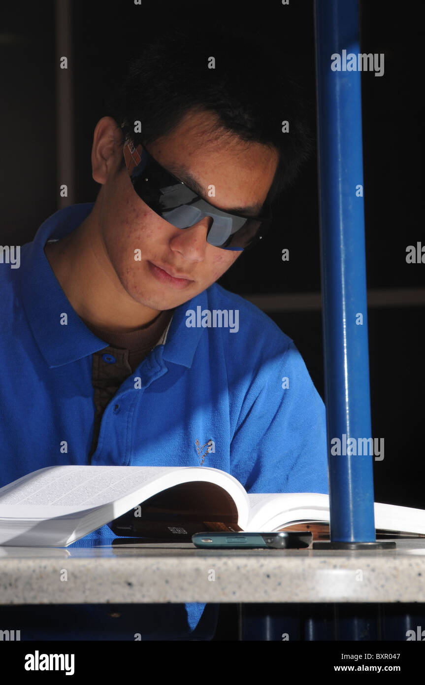 A college student studying outdoors sits in the shade and wears eye ...