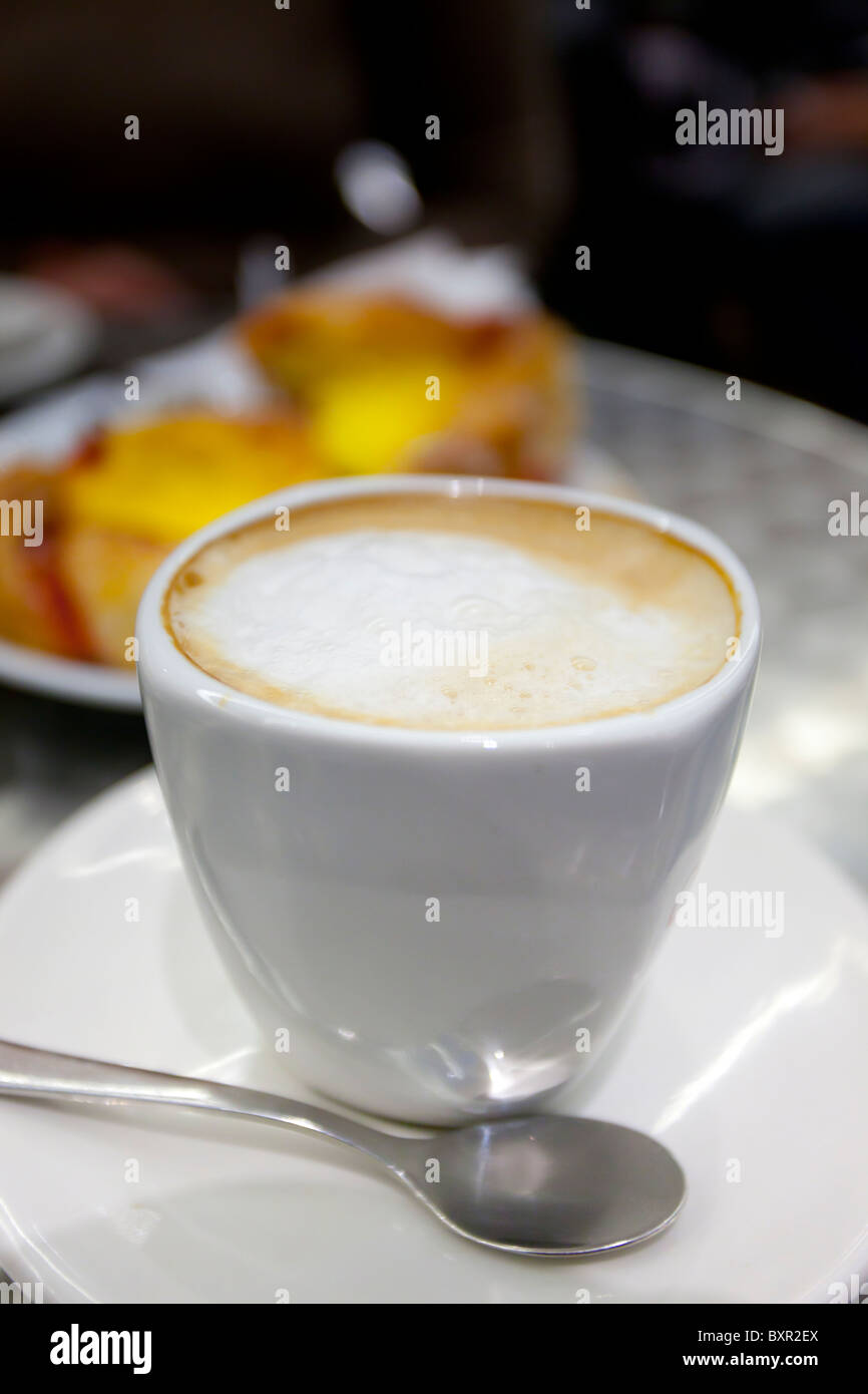 Close-up of a frothy cappuccino in a white cup with a blurred pastry in ...