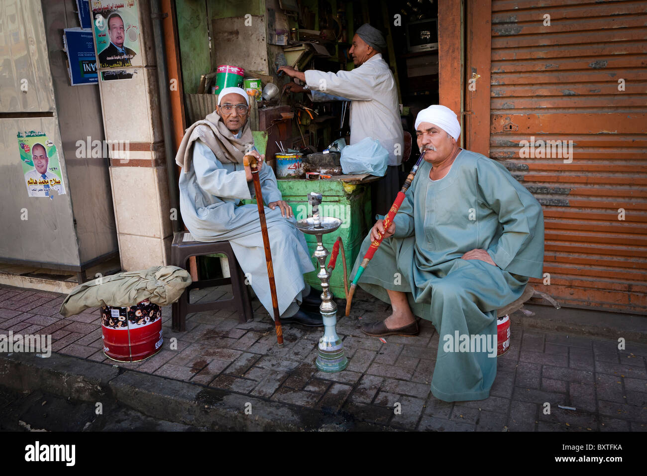 Two men sit relaxing and smoking shisha outside shop as owner works ...