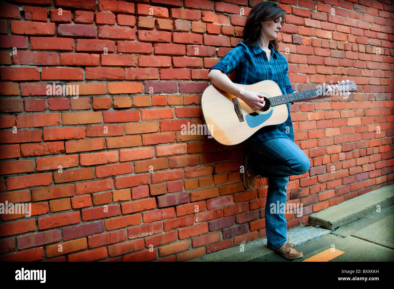Female guitarist playing guitar while leaning against red brick wall ...