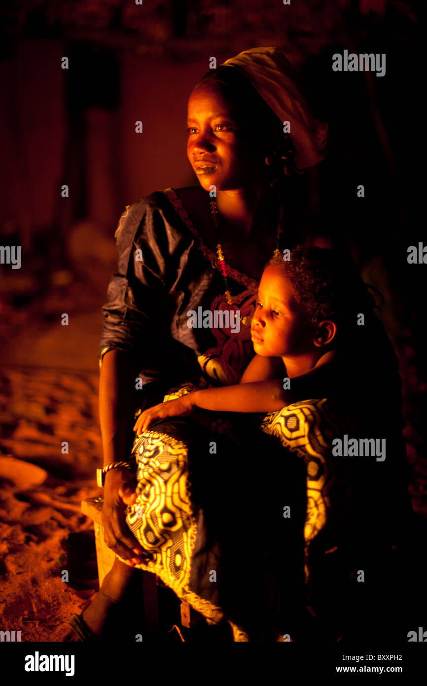 A woman and child sit next to the fire, where a feast grills. Tabaski ...