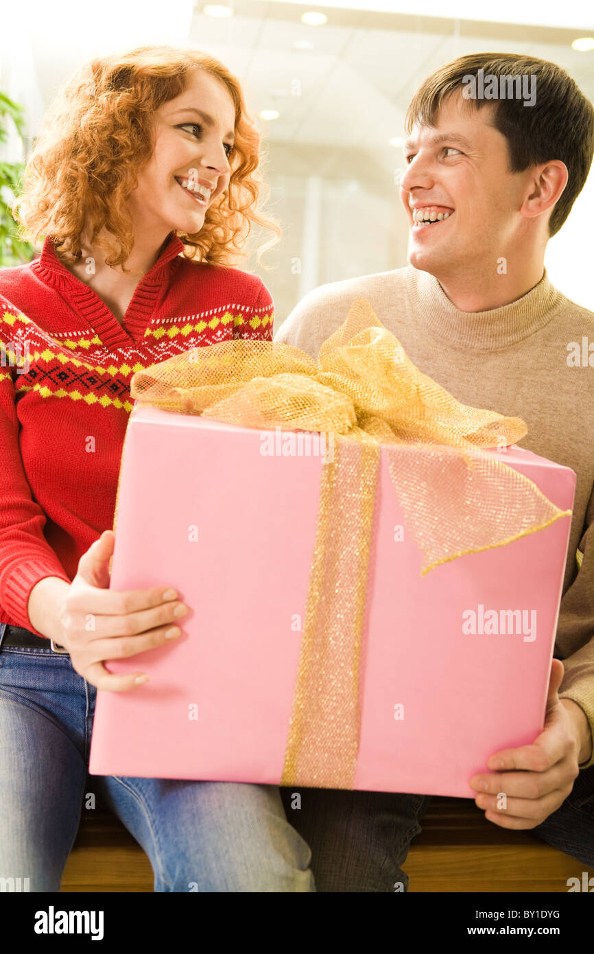 Portrait of smiling couple holding big pink gift box and looking at ...