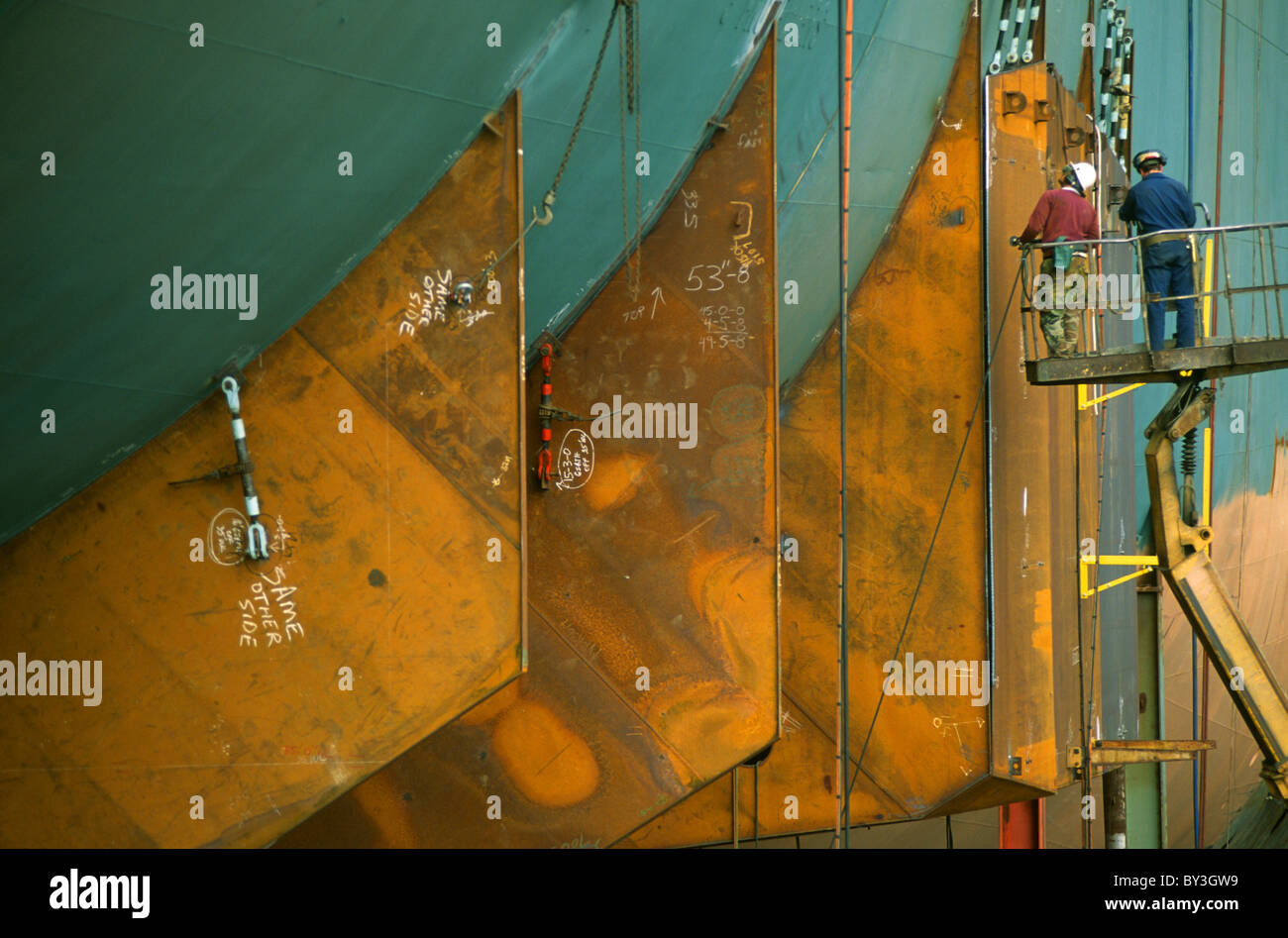 Two workers inspecting outside hull of ship under construction at ...