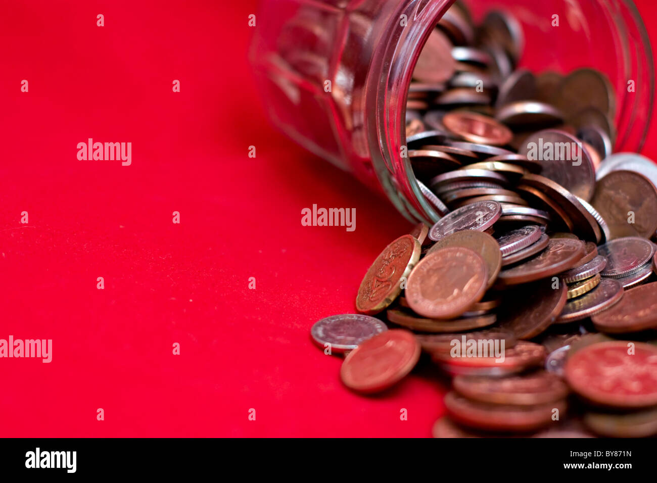 British coins spill out of a plain glass jar against a red background ...