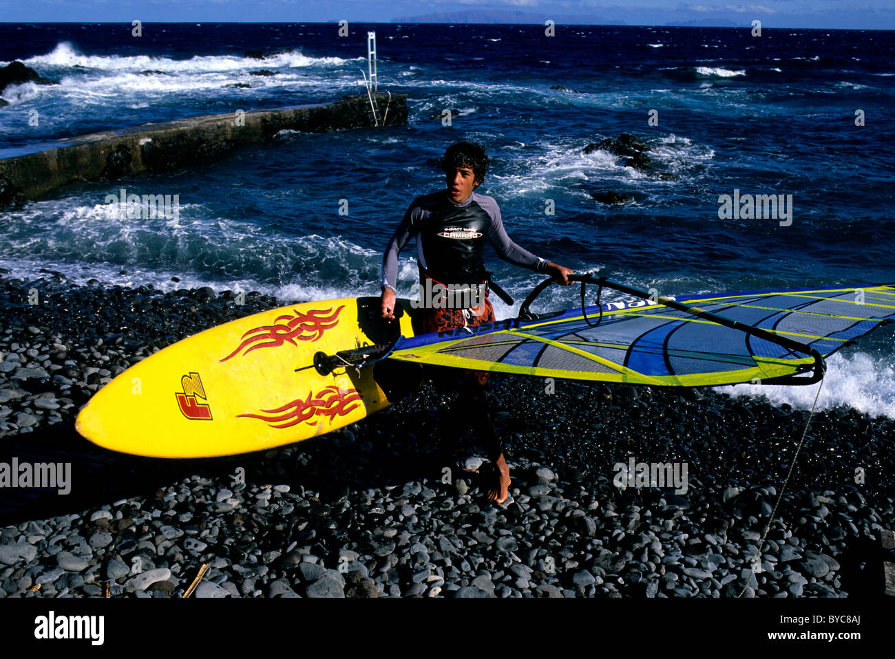 A windsurfer pulling up his board onto a stony beach in Madeira after ...