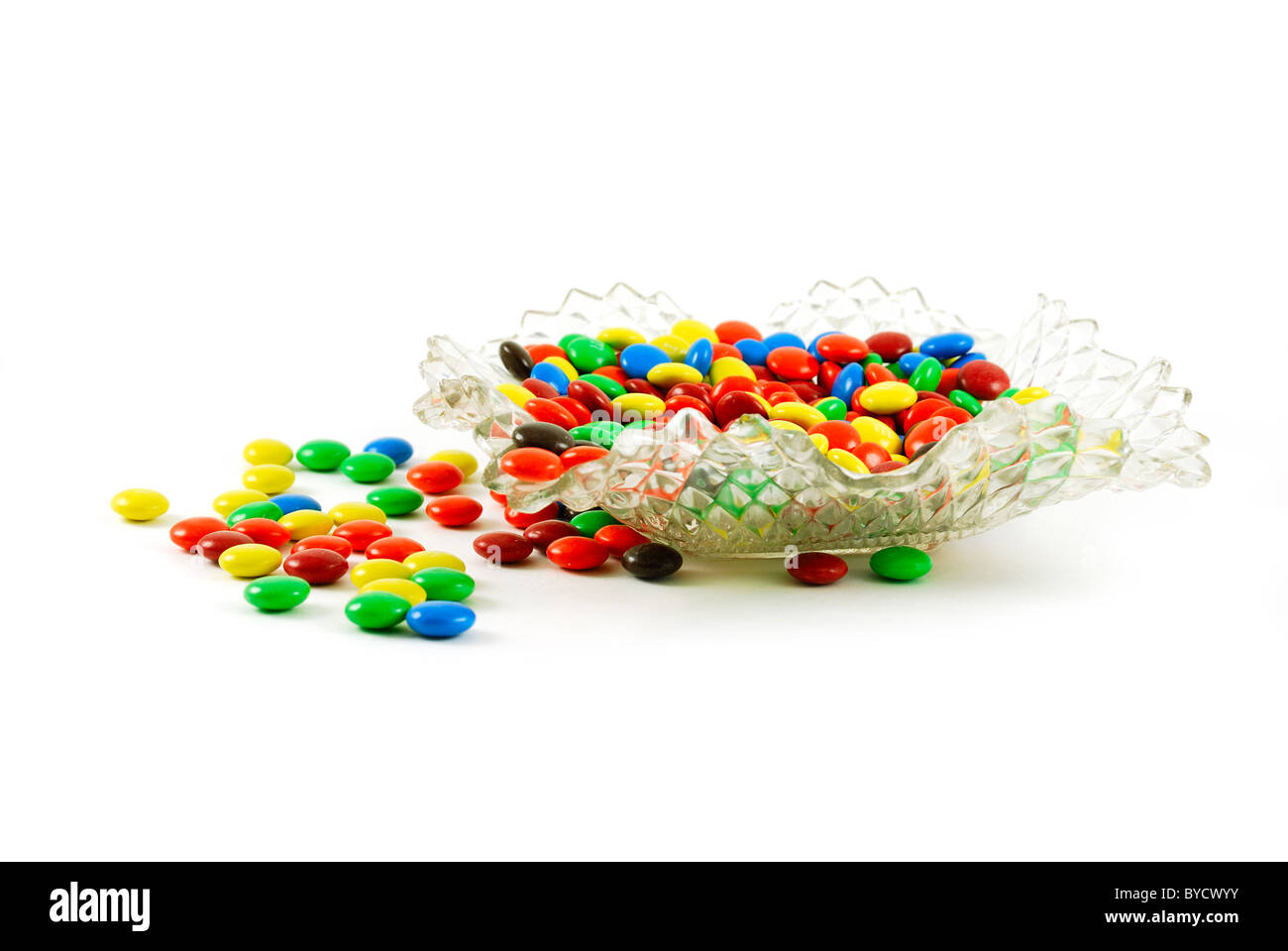 A glass candy dish full of colorful candy with several pieces spilled ...