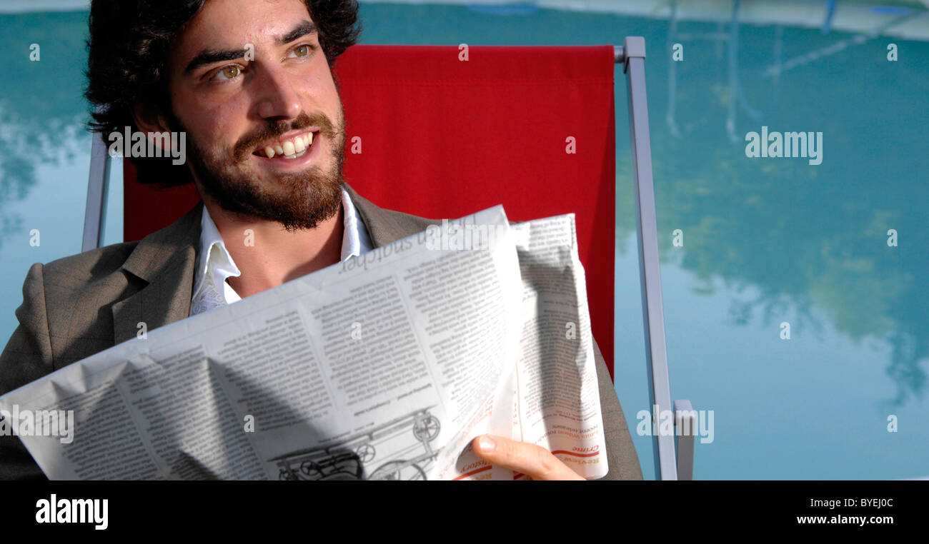 A happy young man smiling and laughing reading paper by swimming pool ...