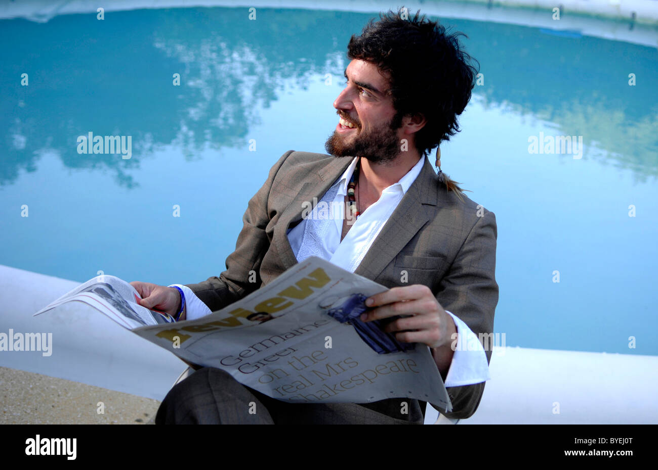 A happy young man smiling and laughing reading paper by swimming pool ...
