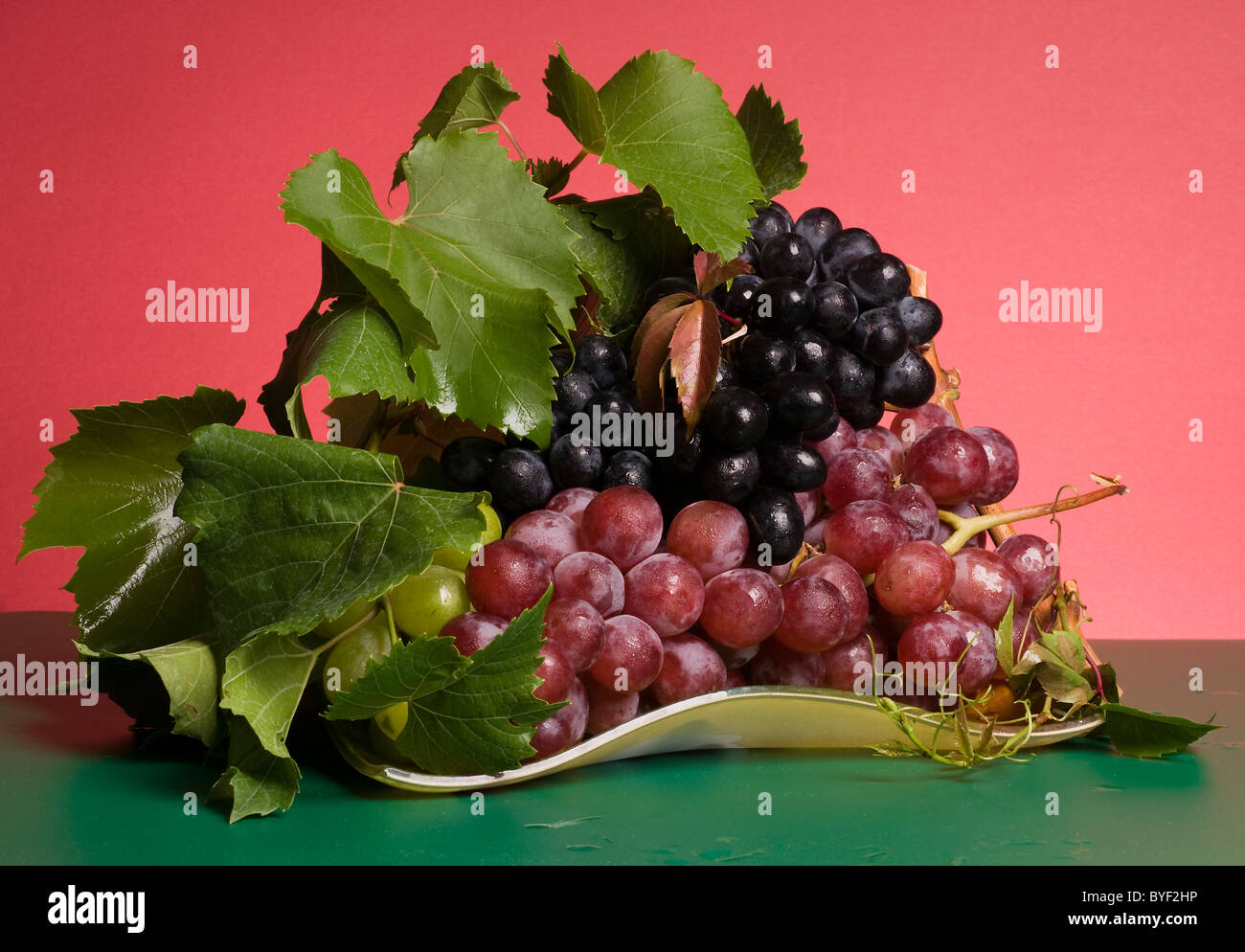 Still-life with multicolor raw grape and leaves over green background ...