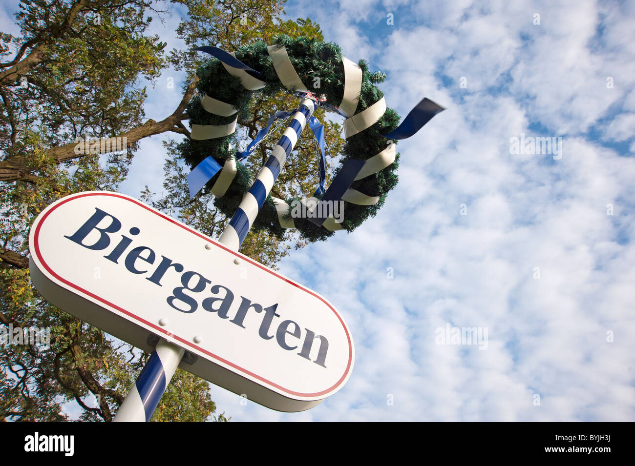 Biergarten sign on a pole in front of blue sky with clouds and a tree ...