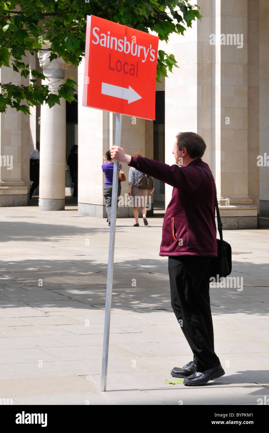 Man holding placard sign pointing to location of a new Sainsburys Local ...