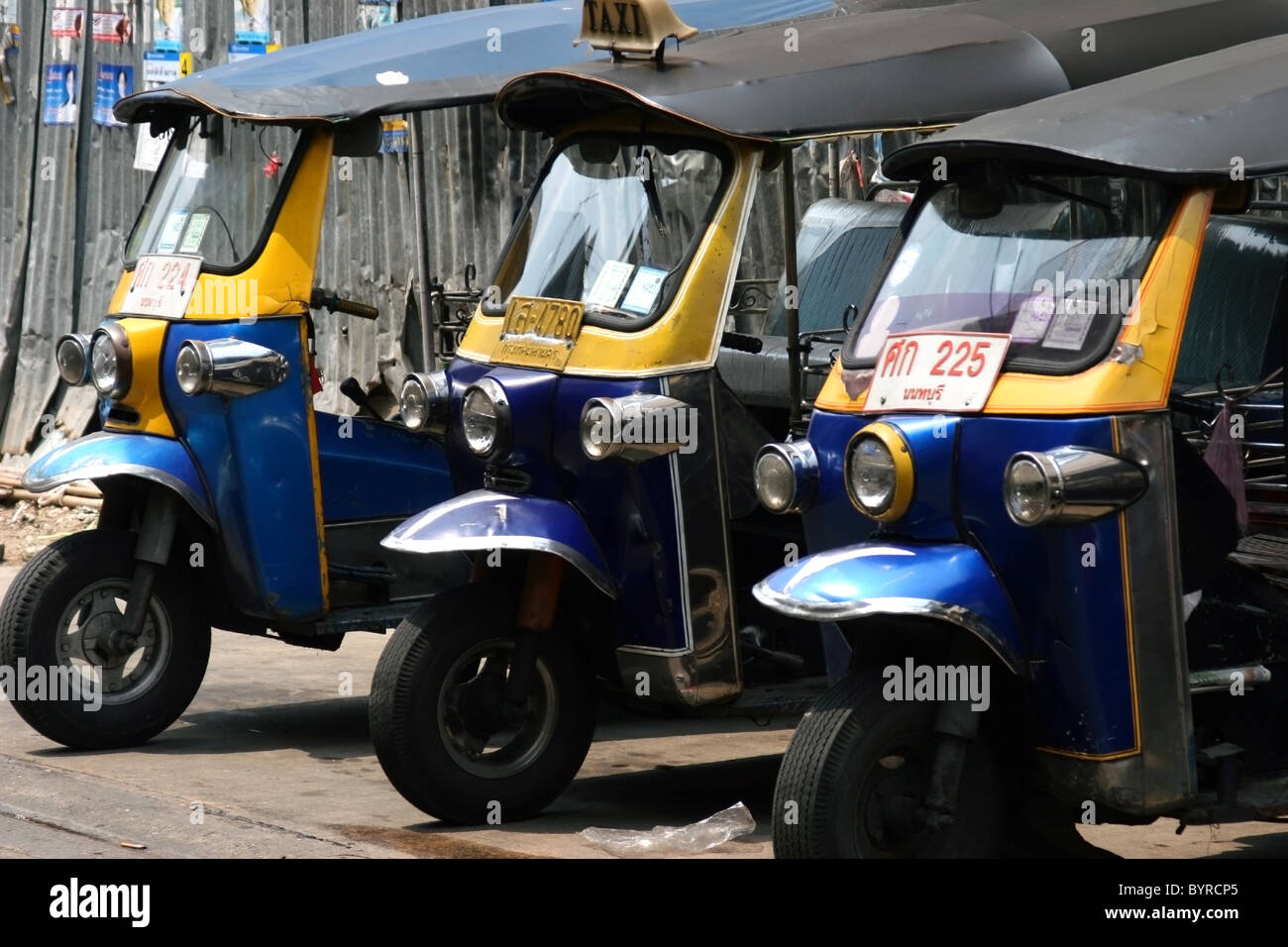 A row of colorful blue and yellow tuk-tuks are parked on a city street in Bangkok, Thailand ...
