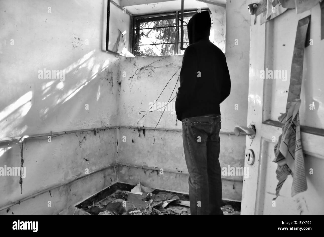 Hooded male figure looking through window of abandoned house. Black and ...