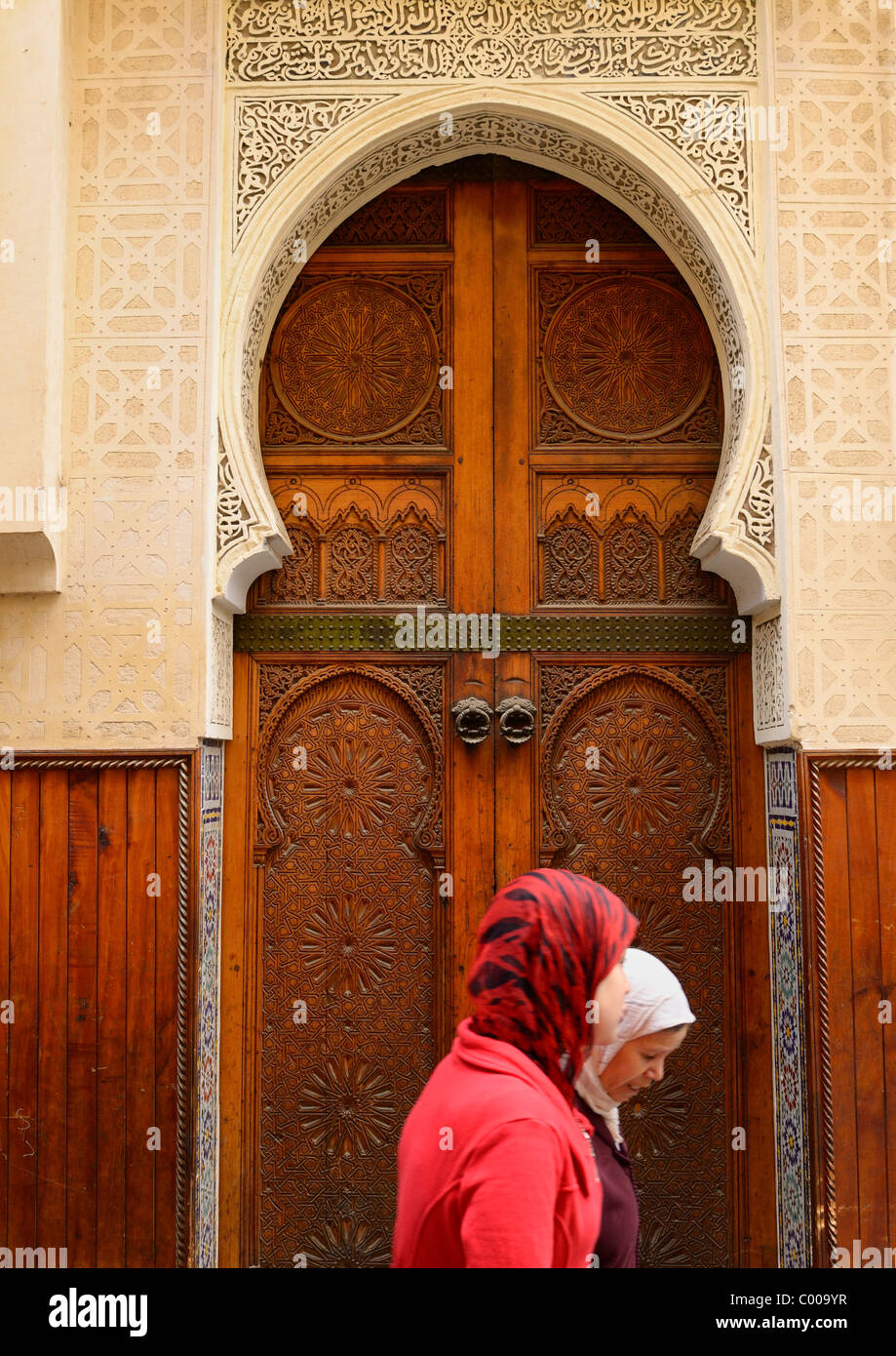 Two women in red hijab passing ornate wooden doors of a Mosque in Fes ...