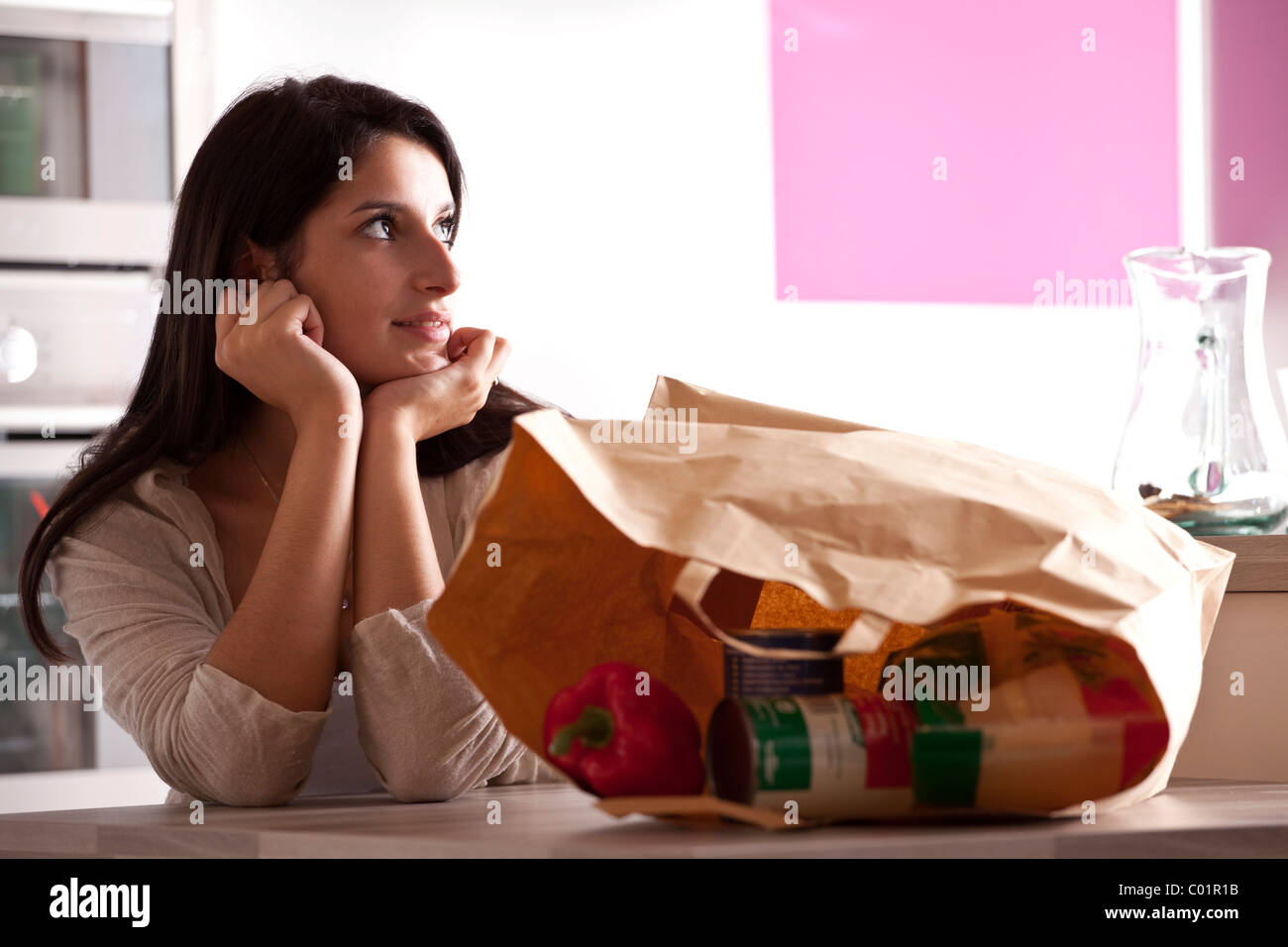Young woman sitting at a dining table in the kitchen, thinking, paper ...