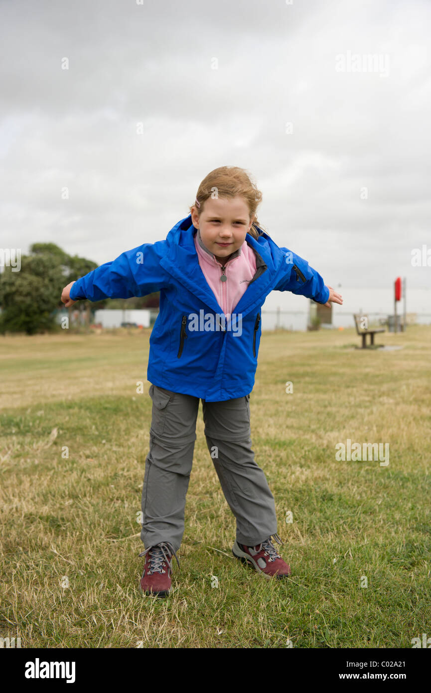 A young girl in outdoor clothes, leans into a high wind with her arms ...