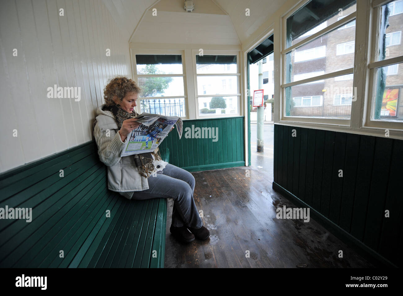 Woman reading local newspaper sitting in wooden bus shelter waiting for ...