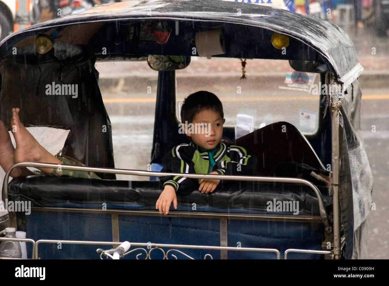 A young boy is gazing outside of a parked tuk tuk on a rainy day on a ...