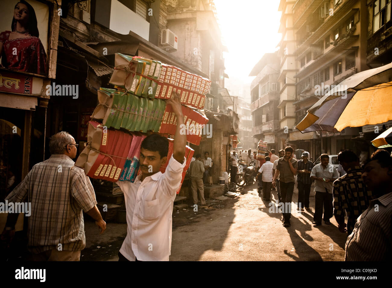 A young man carries a stack of boxes through a crowded, dusty and ...
