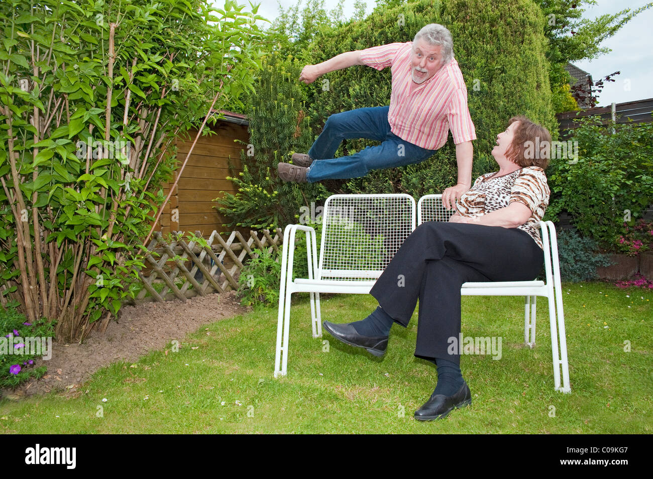 Senior citizen jumping for joy over a bench on which his wife is ...
